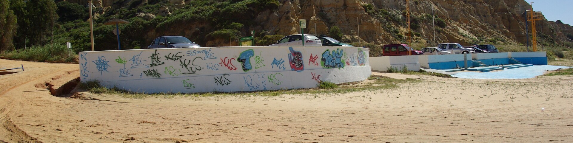 Uma praia arenosa com paredes grafitadas e carros estacionados.