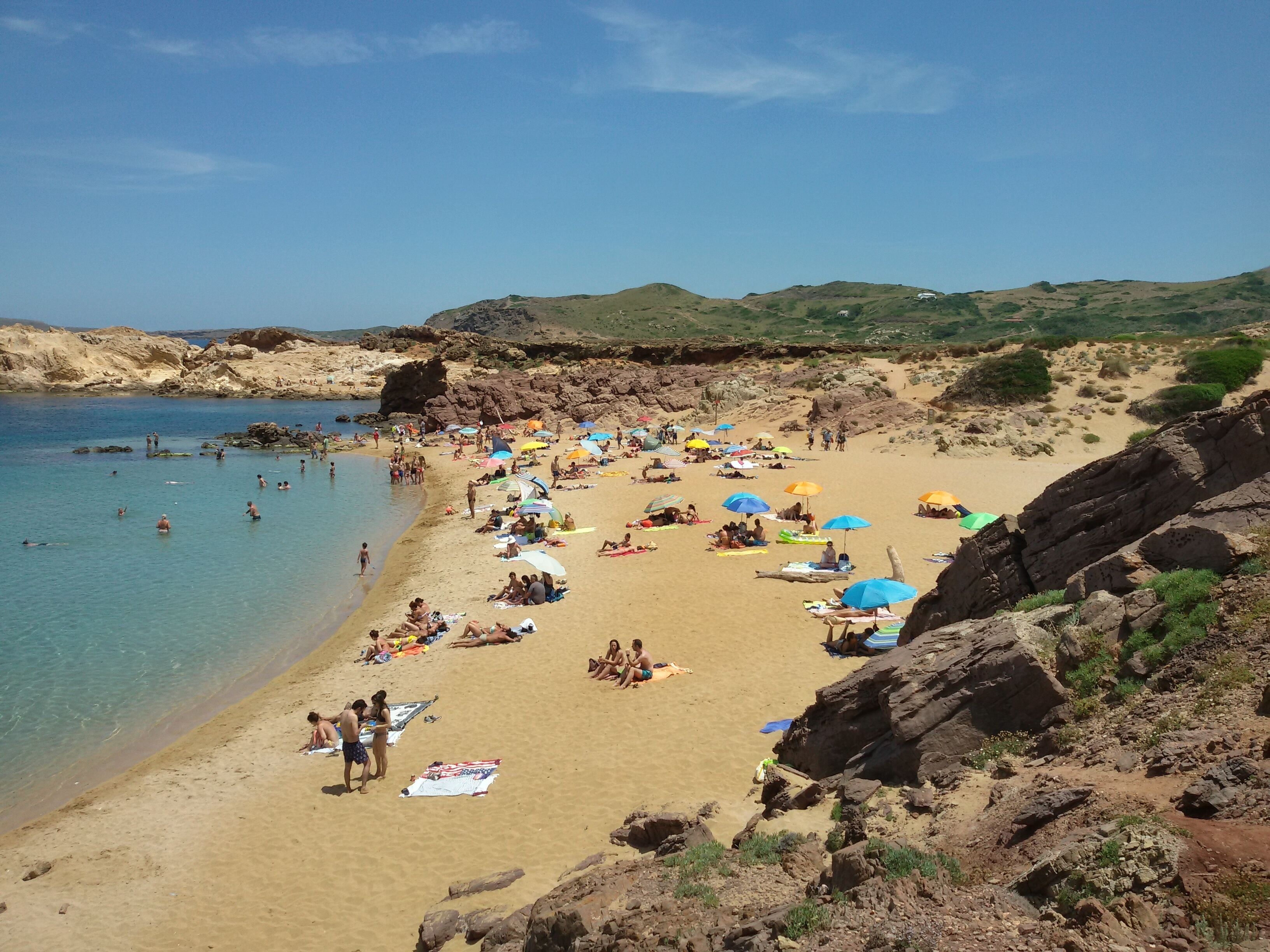 Uma praia movimentada com pessoas tomando sol e nadando, cercada por falésias e colinas rochosas.