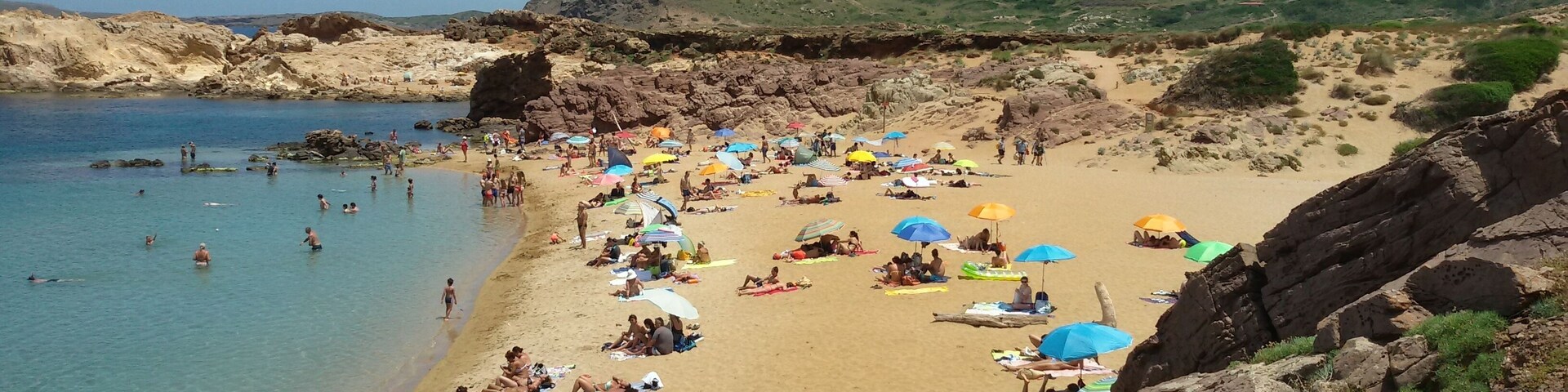 Uma praia movimentada com pessoas tomando sol e nadando, cercada por falésias e colinas rochosas.