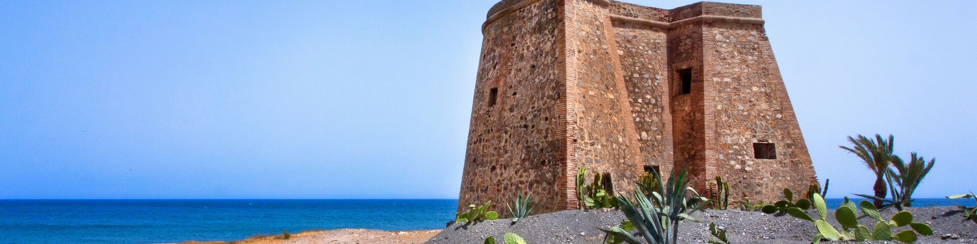 Uma fortaleza costeira com vista ampla para o mar e primeiro plano arenoso com cactos.