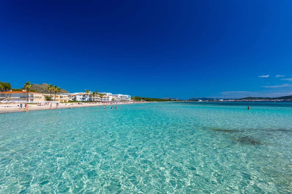 Mar turquesa translúcida, praia de areia e um resort com edifícios brancos e palmeiras.