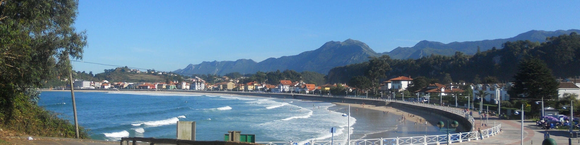 Um calçadão costeiro com cerca de madeira, praia e montanhas ao fundo.