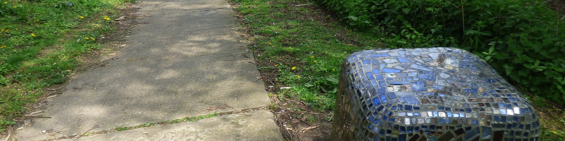 Um caminho de concreto em um parque, com um banco revestido de mosaicos e uma grande árvore.