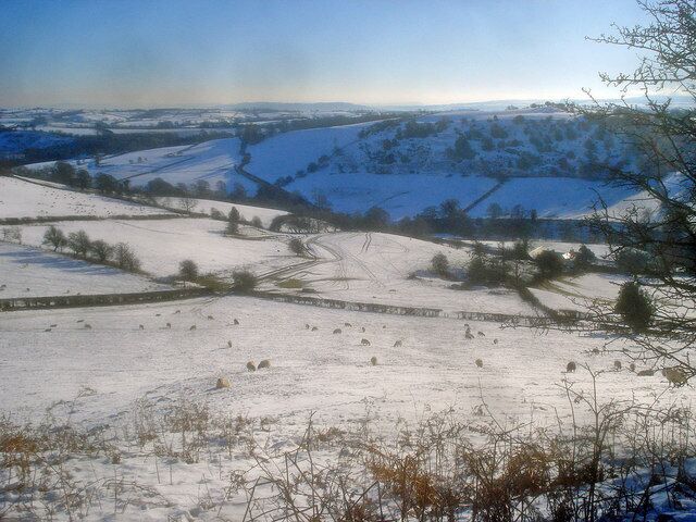 Paisagem rural coberta de neve, com campos, muros de pedra e um caminho sinuoso.