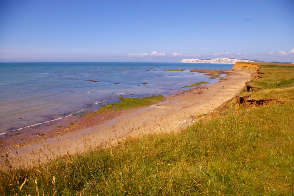 Paisagem costeira com uma praia arenosa, dunas gramadas e falésias brancas ao fundo.