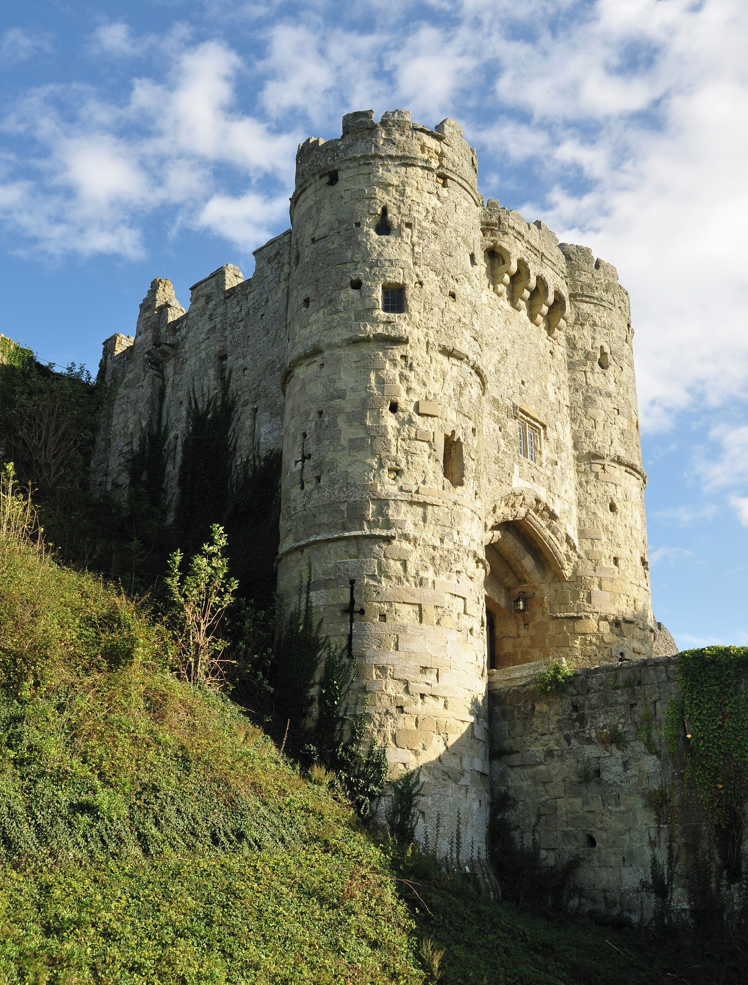 Um castelo medieval com uma torre redonda, rodeado por vegetação e situado contra um c éu claro.