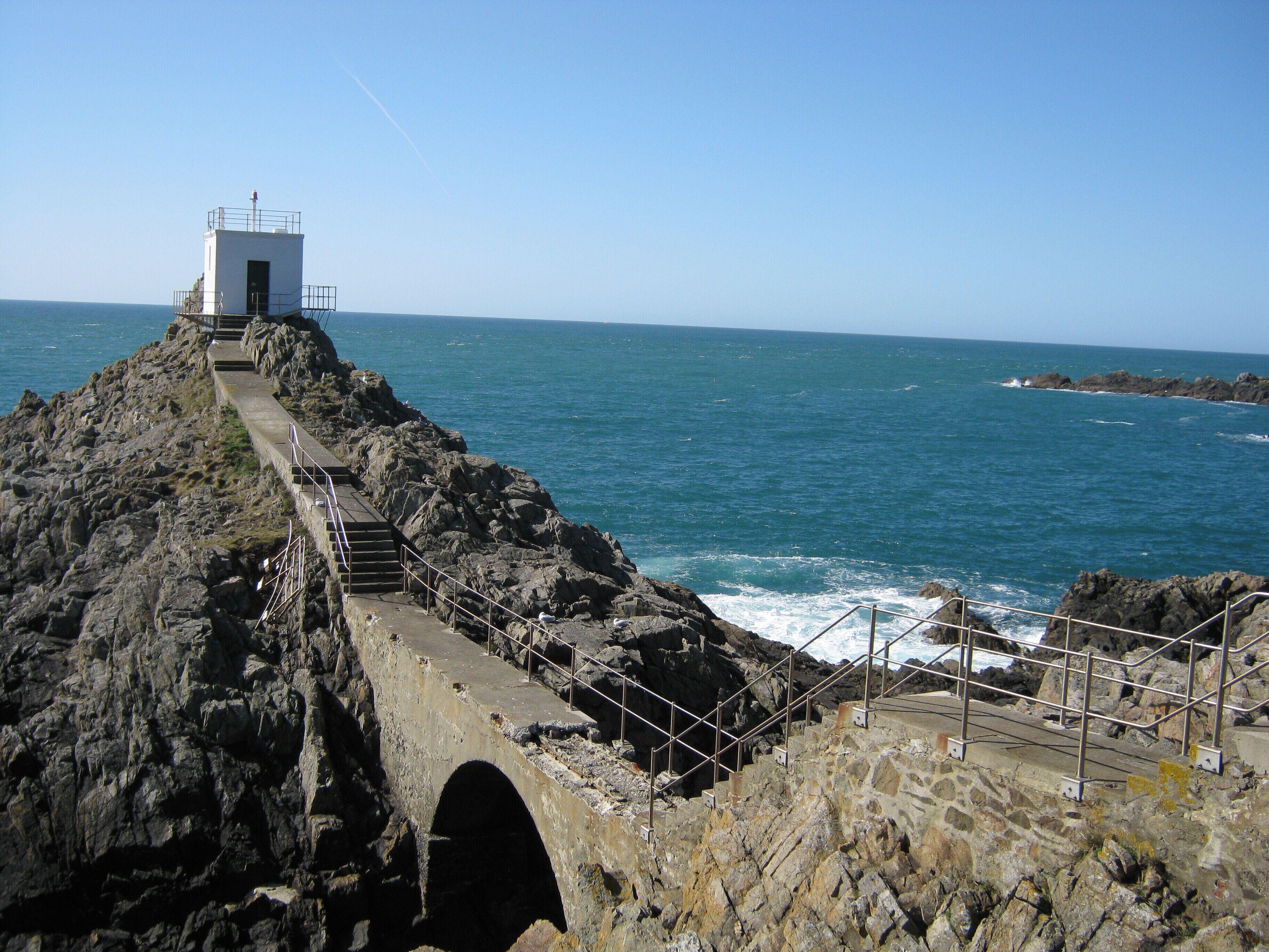 Uma trilha costeira rochosa que leva a um pequeno edifício branco com uma bandeira no topo, com vista para o mar.