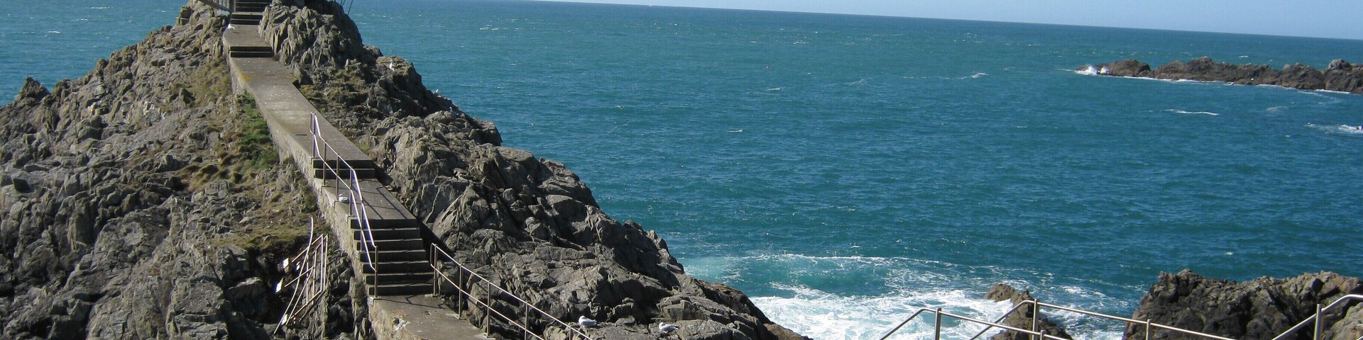 Uma trilha costeira rochosa que leva a um pequeno edifício branco com uma bandeira no topo, com vista para o mar.