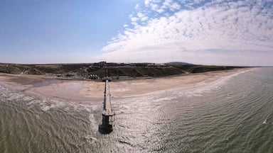 Vista aérea de uma praia com um cais que se estende para o mar, uma faixa de areia e uma colina distante com edifícios.