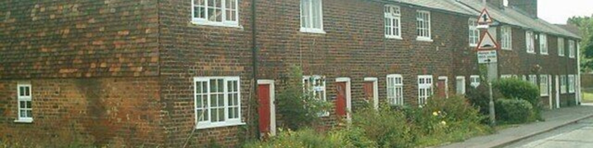 Fileira de casas de tijolo vermelho com janelas e portas brancas, uma rua com calçada e uma placa de aviso triangular.
