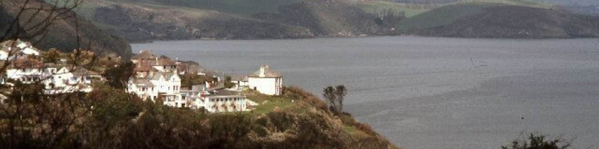 Uma vila costeira com edifícios brancos, uma vasta extensão de água e colinas suaves ao fundo.