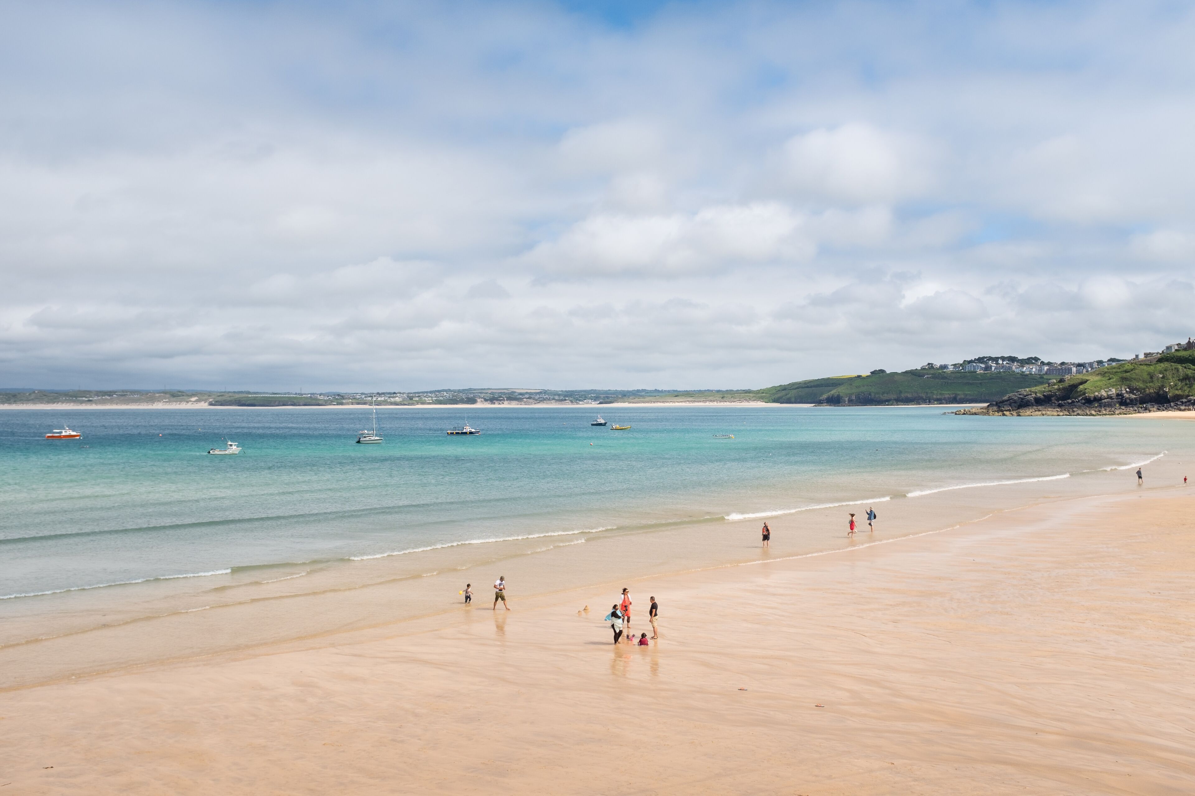 Uma praia arenosa com águas calmas, barcos e uma colina distante com edifícios.