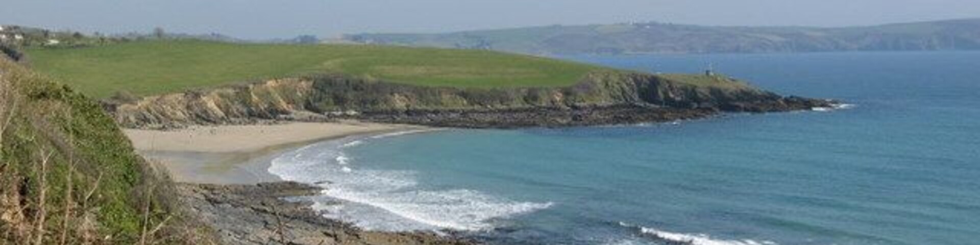 Paisagem costeira com uma praia arenosa, litoral rochoso e colinas gramadas.
