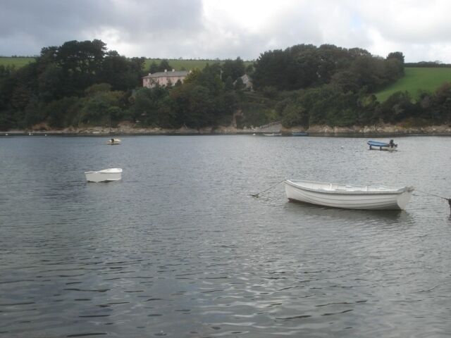 Um lago tranquilo com duas embarcações ancoradas, cercado por árvores e um edifício distante.