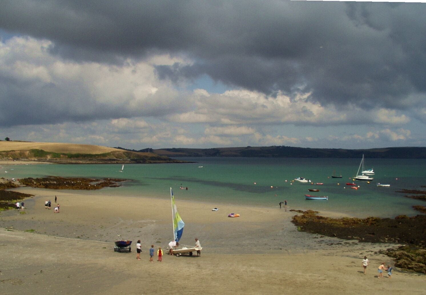 Uma praia com barcos, pessoas e um veleiro na orla.