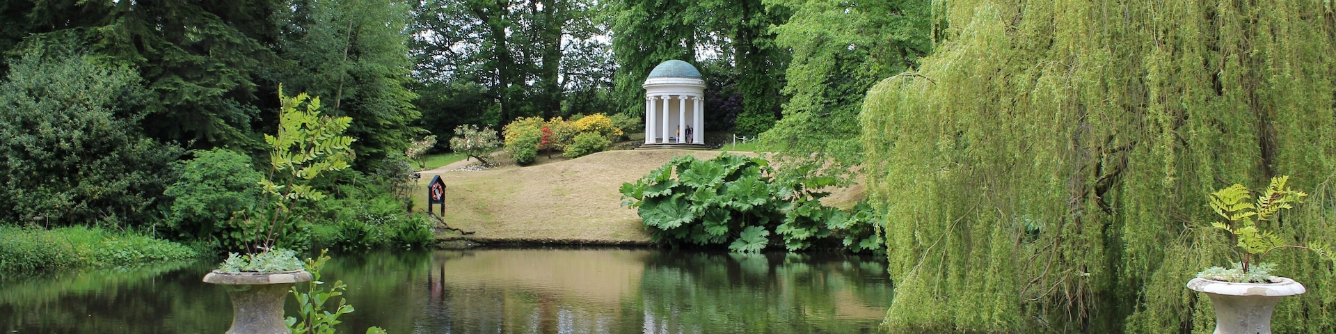 Um jardim tranquilo com lago, um gazebo branco, vegetação exuberante e um salgueiro-chorão.