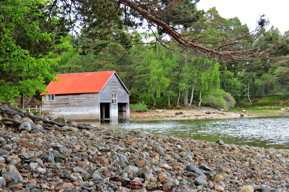 Uma cabana de madeira com telhado vermelho, à beira de uma costa rochosa e margem de rio arborizada.