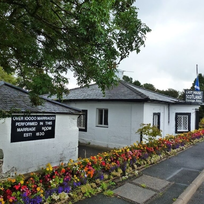 Um edifício branco com uma placa indicando 'LAST HOUSE SCOTLAND' e outra placa que informa 'MAIS DE 10000 CASAMENTOS CELEBRADOS NESTA SALA DE CASAMENTOS, FUNDADA EM 1830'.