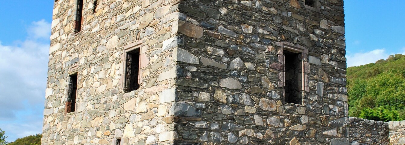 Um edifício de pedra com telhado inclinado, rodeado por uma área gramada e uma cerca de arame.