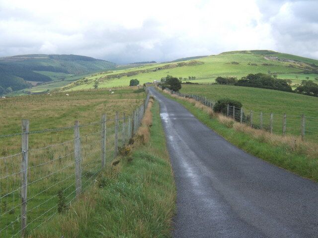 Uma estrada rural estreita ladeada por campos gramados e uma cerca, serpenteando por uma paisagem montanhosa.