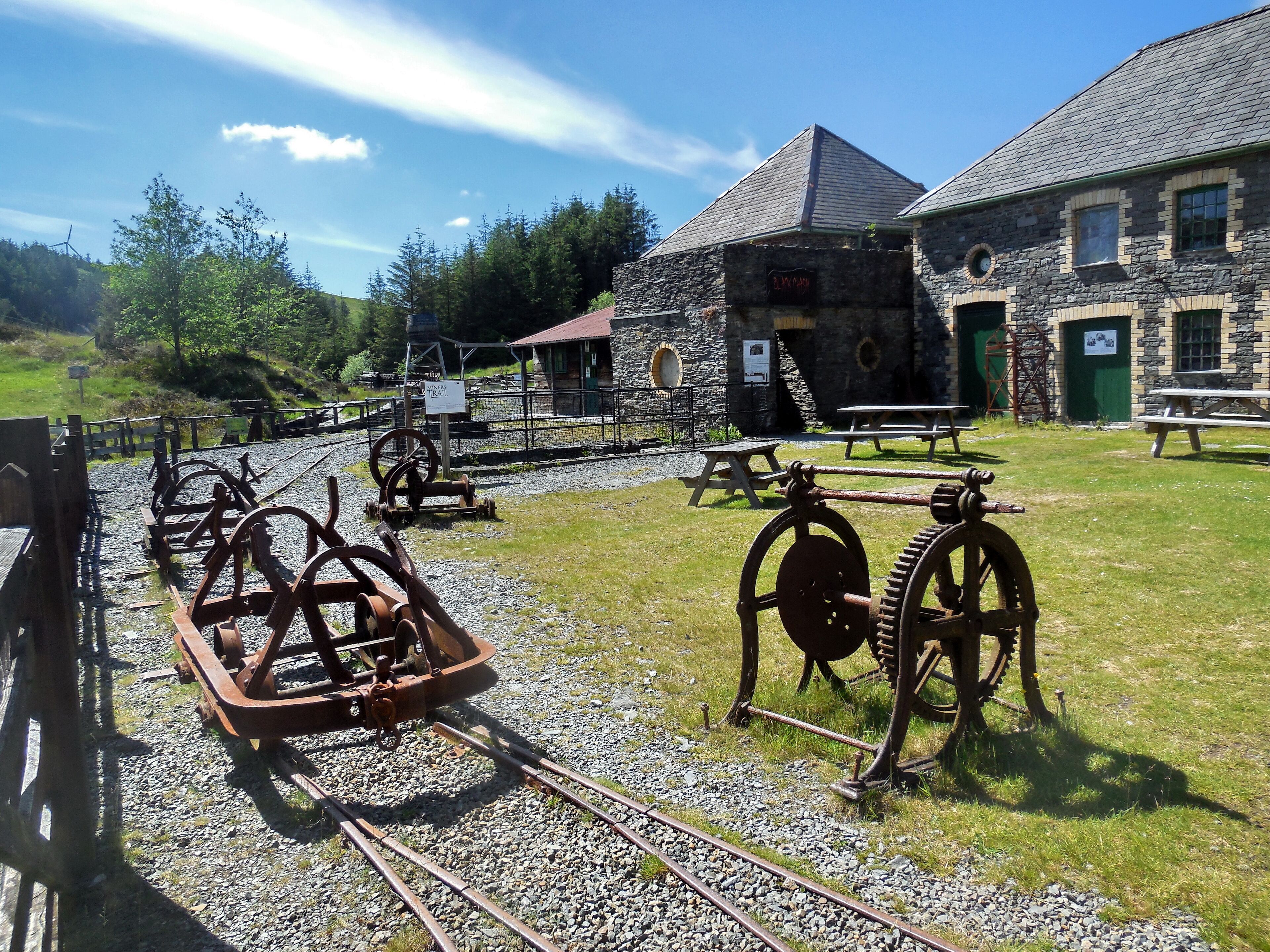 Sítio histórico com equipamentos ferroviários antigos, edifícios de pedra, mesas de piquenique e uma placa indicando a 'Trilha'.