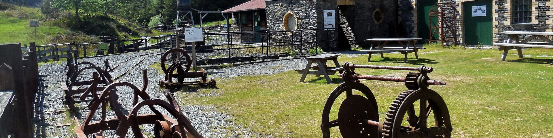 Sítio histórico com equipamentos ferroviários antigos, edifícios de pedra, mesas de piquenique e uma placa indicando a 'Trilha'.