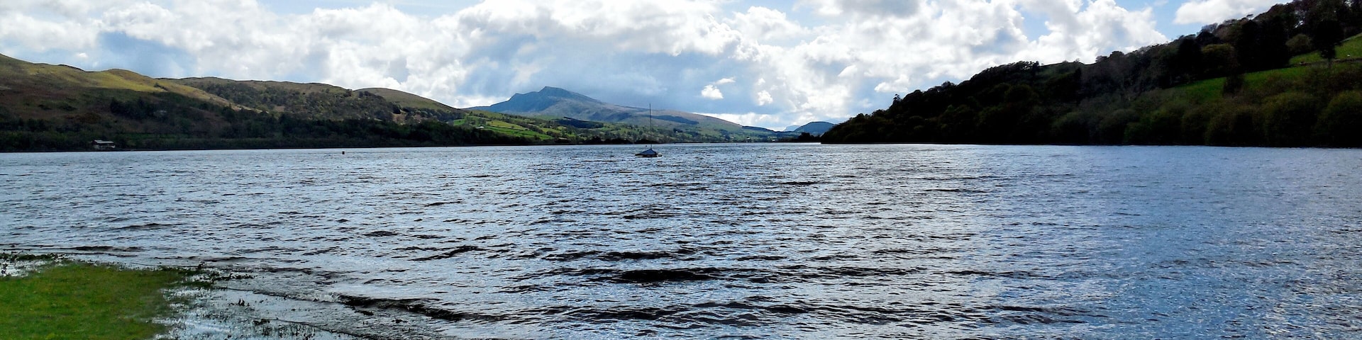 Um lago com um veleiro, cercado por colinas e um céu parcialmente nublado.