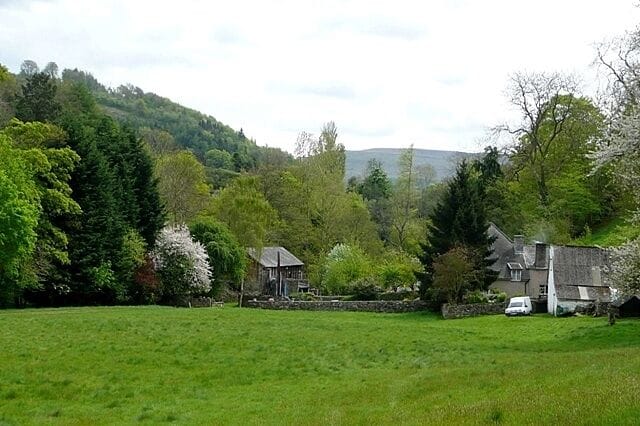 Paisagem rural com um campo verde, um muro de pedra e uma pequena casa rodeada de árvores.