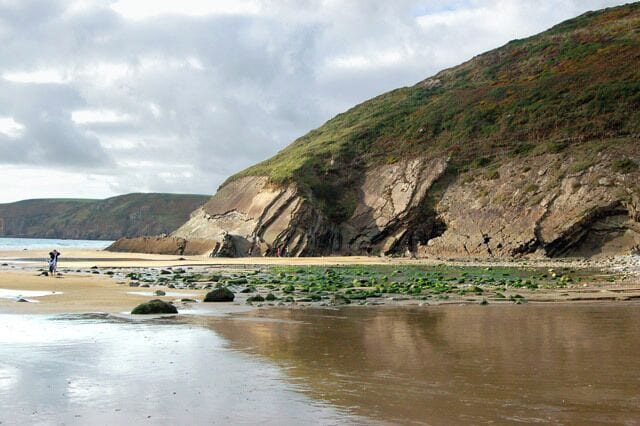 Uma praia costeira com afloramentos rochosos e uma pessoa caminhando pela orla.