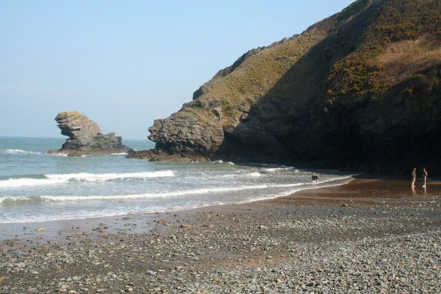 Uma praia rochosa com ondas, um penhasco e duas pessoas caminhando.