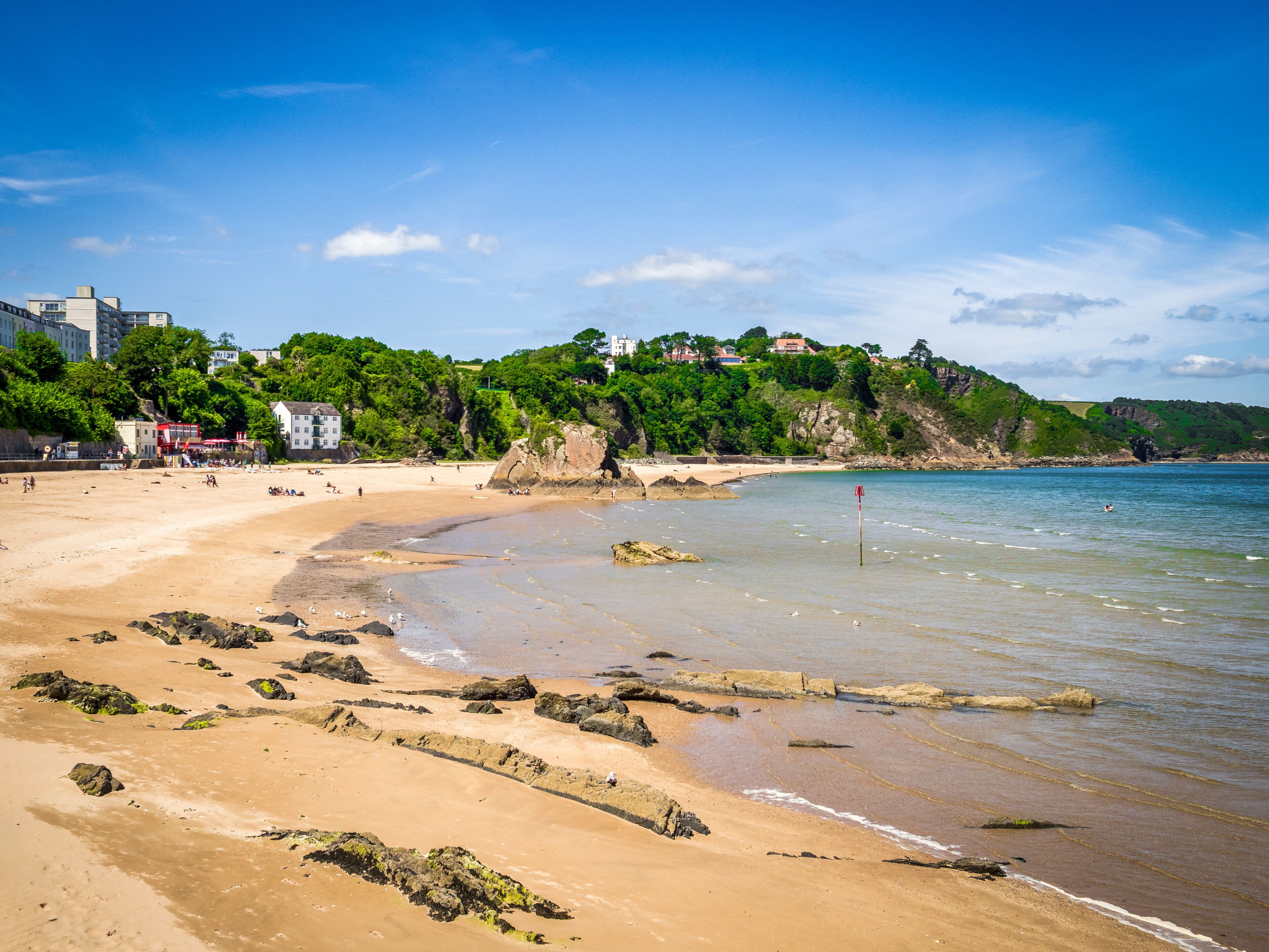 Uma praia arenosa com afloramentos rochosos, água azul cristalina e edifícios numa encosta ao fundo.