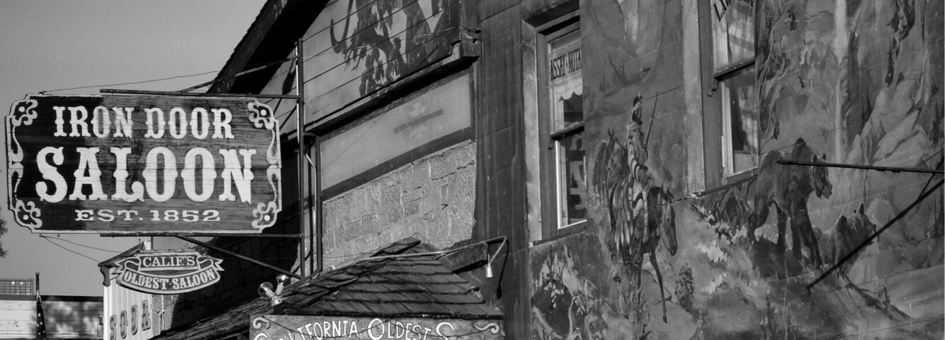 Fotografia a preto e branco de um edifício histórico com placas que indicam 'Iron Door Saloon' e 'California Oldest Saloon'.
