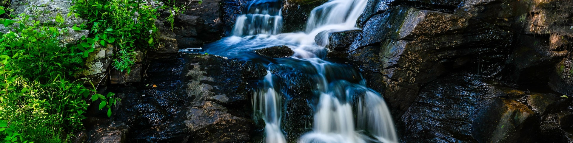 Uma cascata em vários níveis desaguando sobre rochas escuras, cercada por vegetação exuberante.