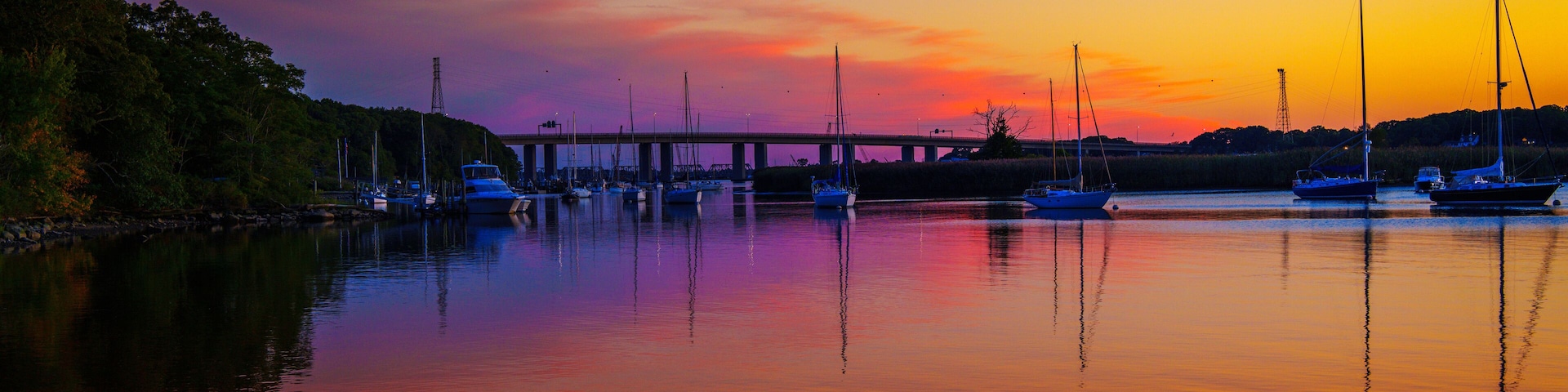 Um pôr do sol sobre um porto tranquilo, com barcos e uma ponte ao fundo.