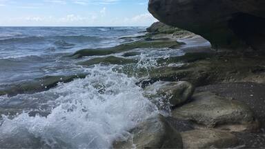 Uma costa rochosa com ondas quebrando na praia e uma grande formação rochosa se projetando na água.