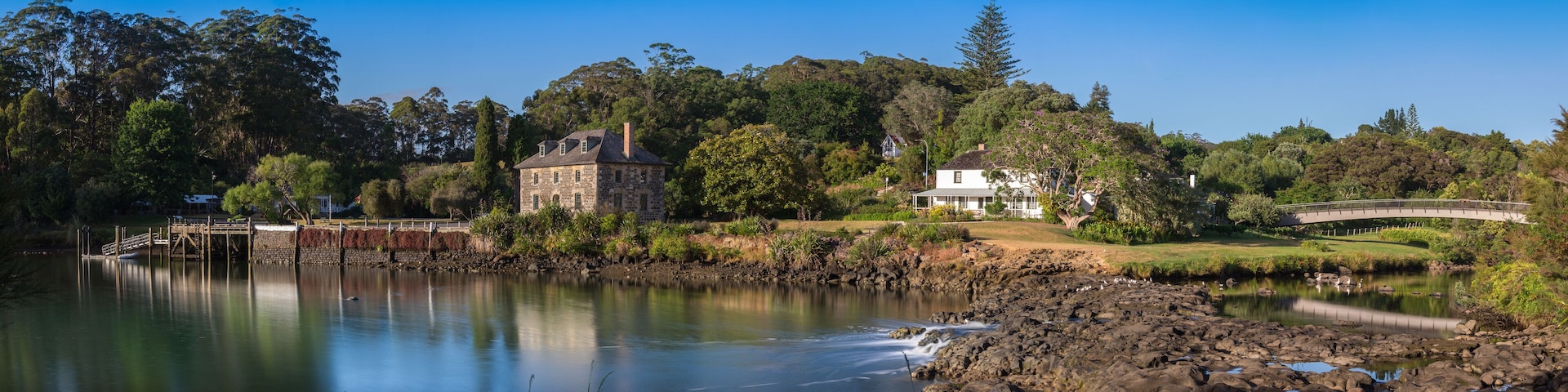 Um lago com uma ponte de pedra, uma casa de pedra e uma casa branca, tudo rodeado por árvores.