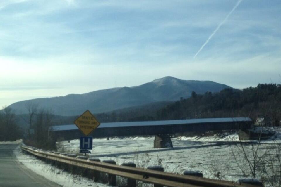 Uma autoestrada com paisagem coberta de neve, uma ponte e um aviso de declive acentuado.