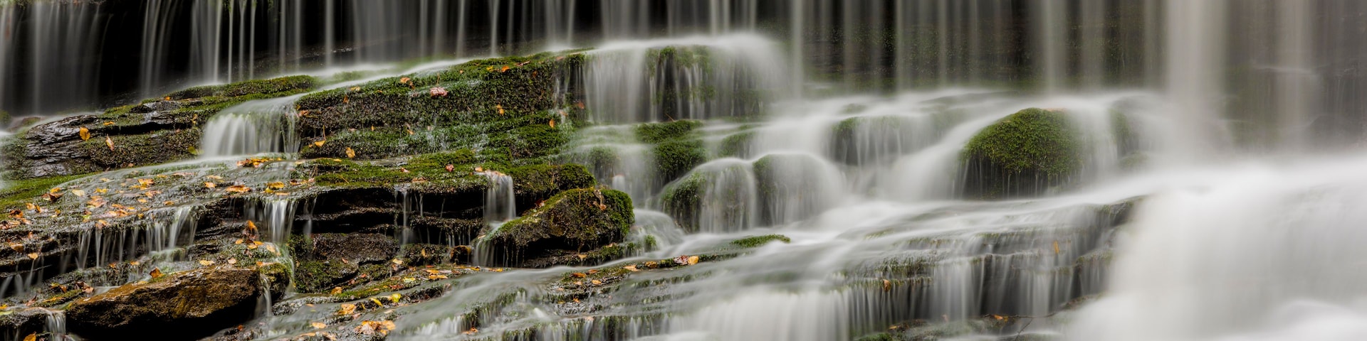 Uma cascata despencando sobre rochas cobertas de musgo, com uma paisagem florestal ao fundo.