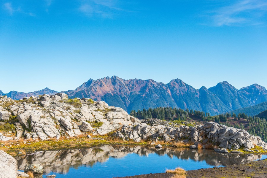 Uma cordilheira com céu azul límpido, terreno rochoso e um lago tranquilo refletindo as montanhas.