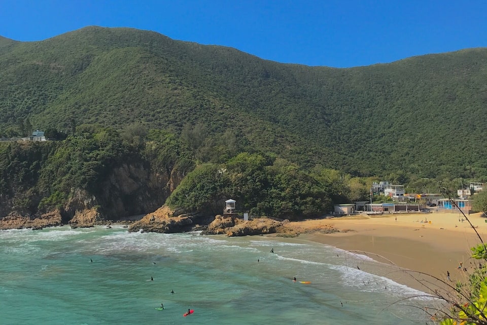Paisagem costeira com uma praia, águas turquesas cristalinas e uma colina arborizada ao fundo.