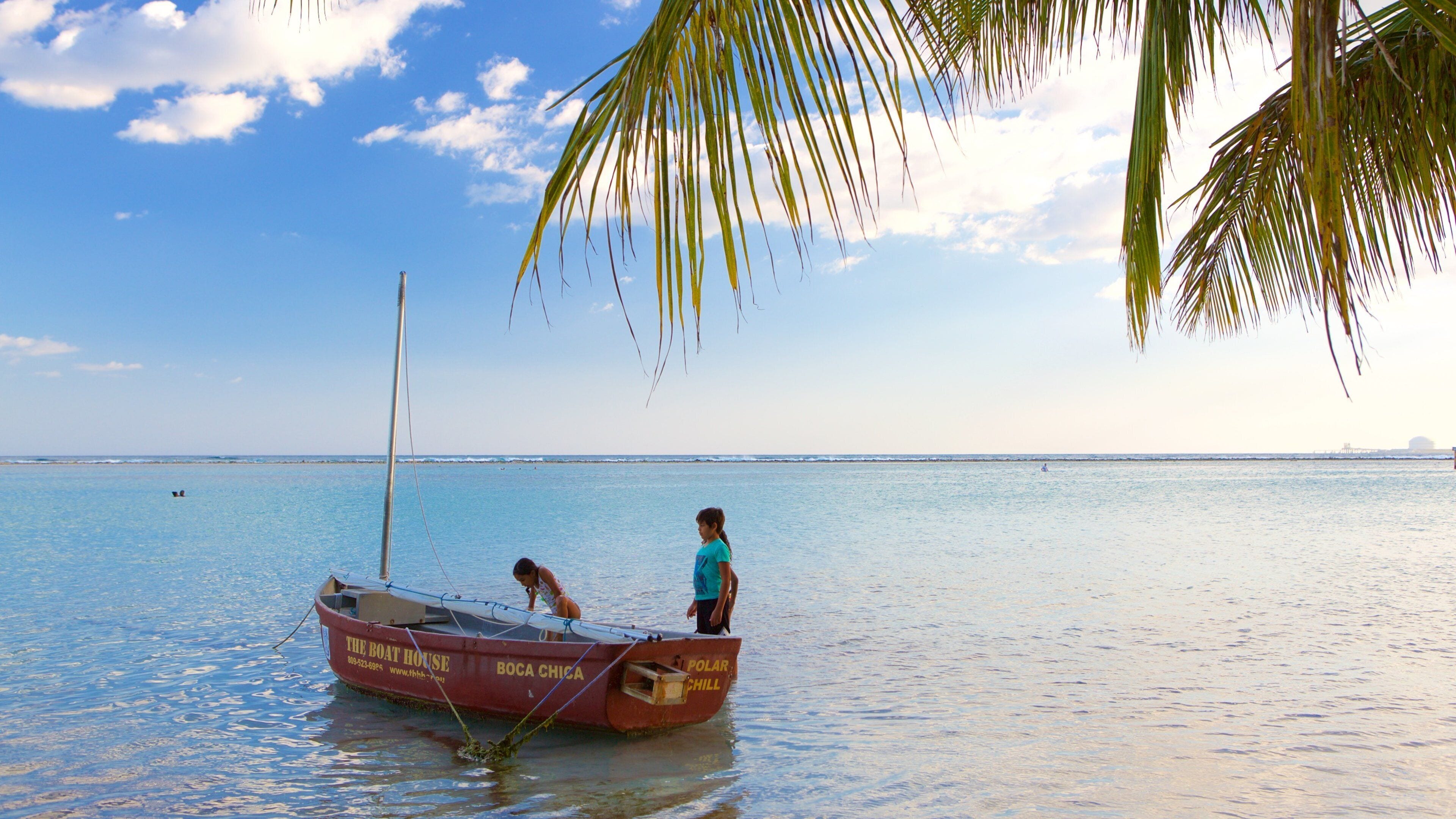 Um barco chamado "The Boat House" com a inscrição "Boca Chica" e "Polar Chill", atracado em um mar calmo com céu claro e palmeiras ao fundo.