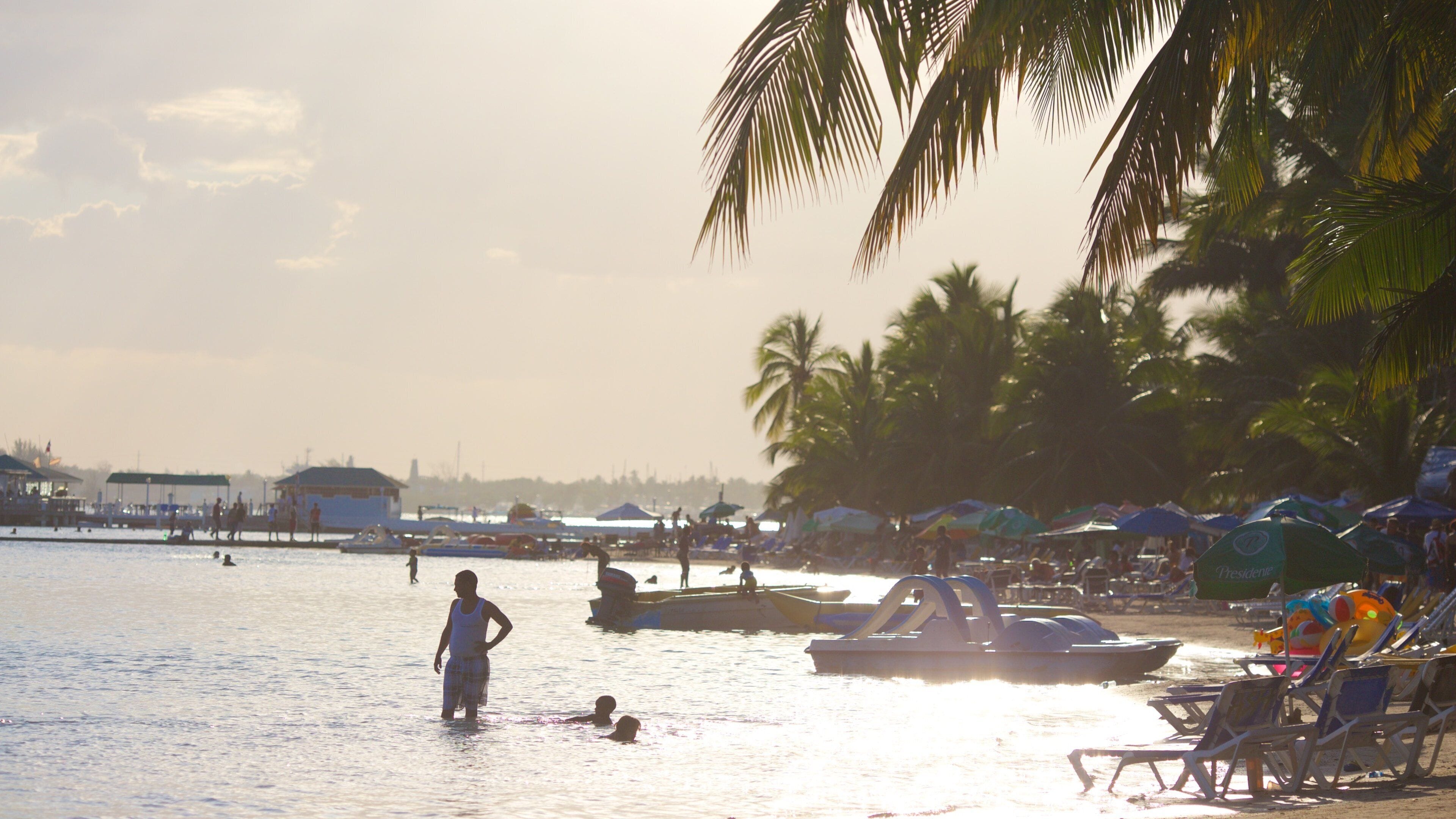 Uma praia com palmeiras, barcos e pessoas nadando.