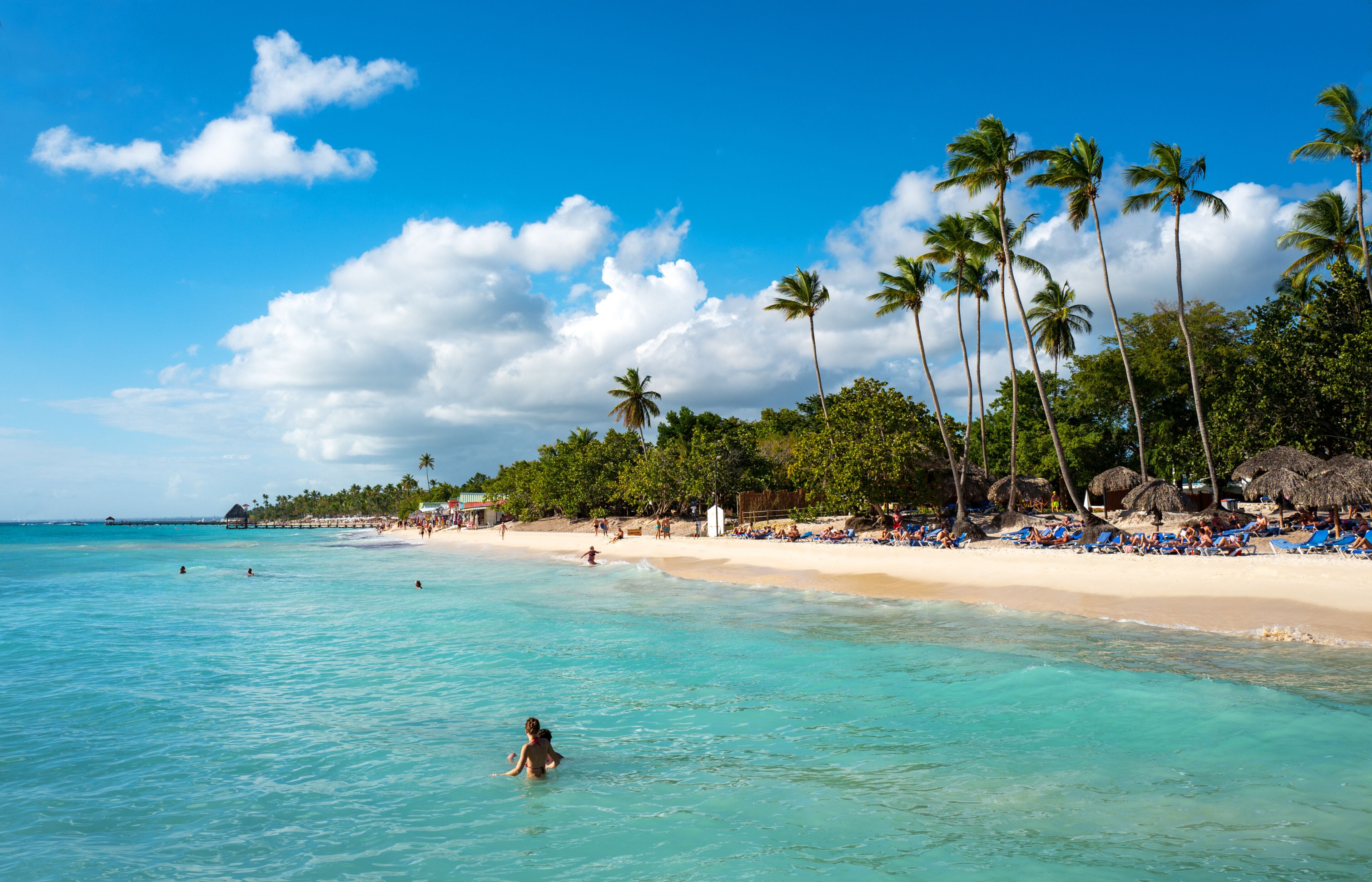 Uma praia tropical com águas turquesa cristalinas, palmeiras e um píer ao longe.