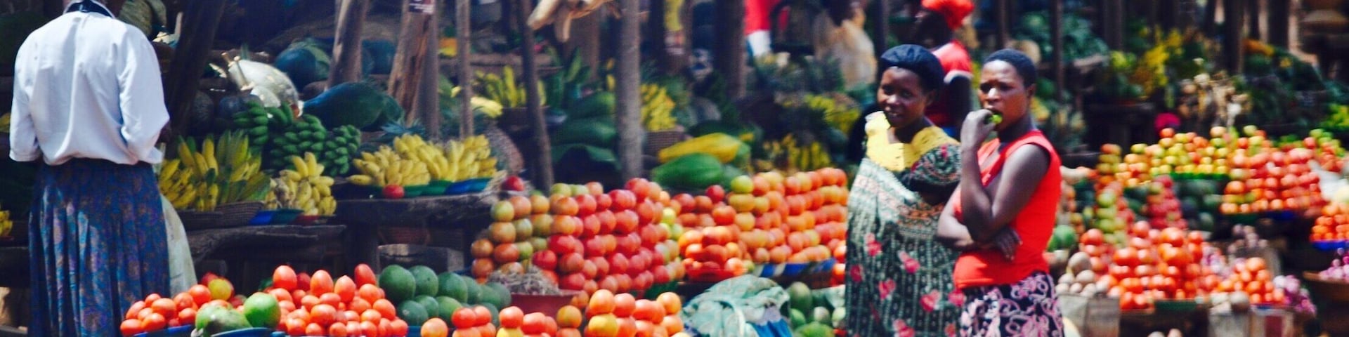 Um mercado ao ar livre movimentado, com várias frutas e legumes expostos em mesas de madeira e pendurados no telhado de palha.