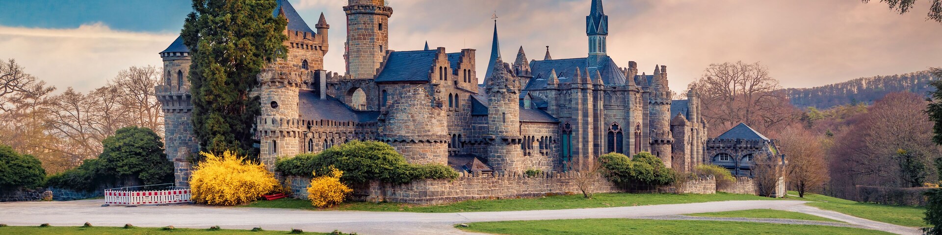Um castelo imponente com múltiplas torres e ameias, cercado por um gramado bem cuidado e árvores.