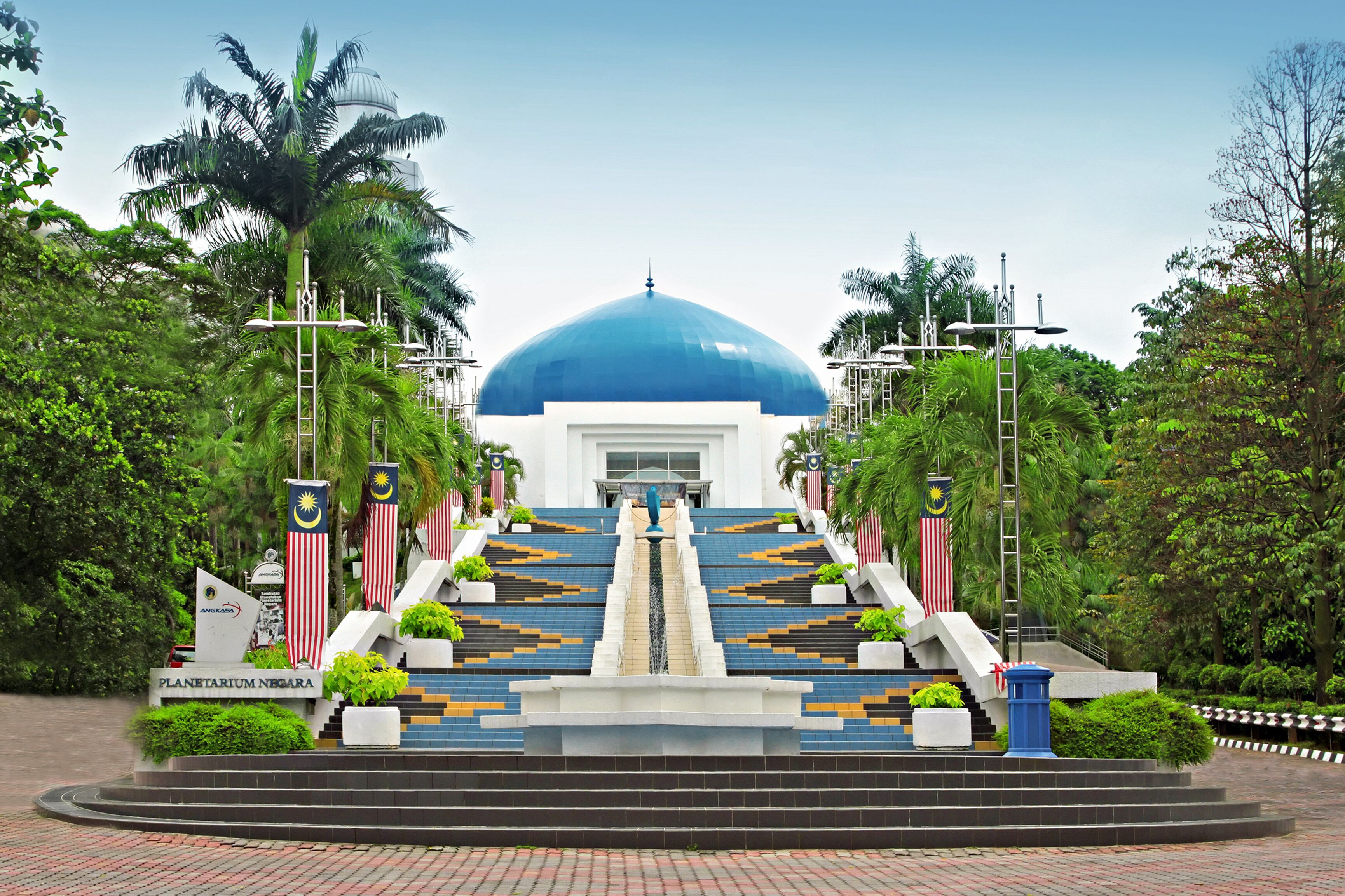 Uma escadaria imponente leva a um edifício grande com cúpula azul, cercado por vegetação exuberante e bandeiras da Malásia.