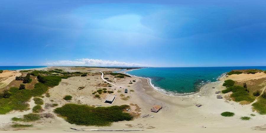 Paisagem costeira com uma praia arenosa, um caminho sinuoso e algumas construções espalhadas.