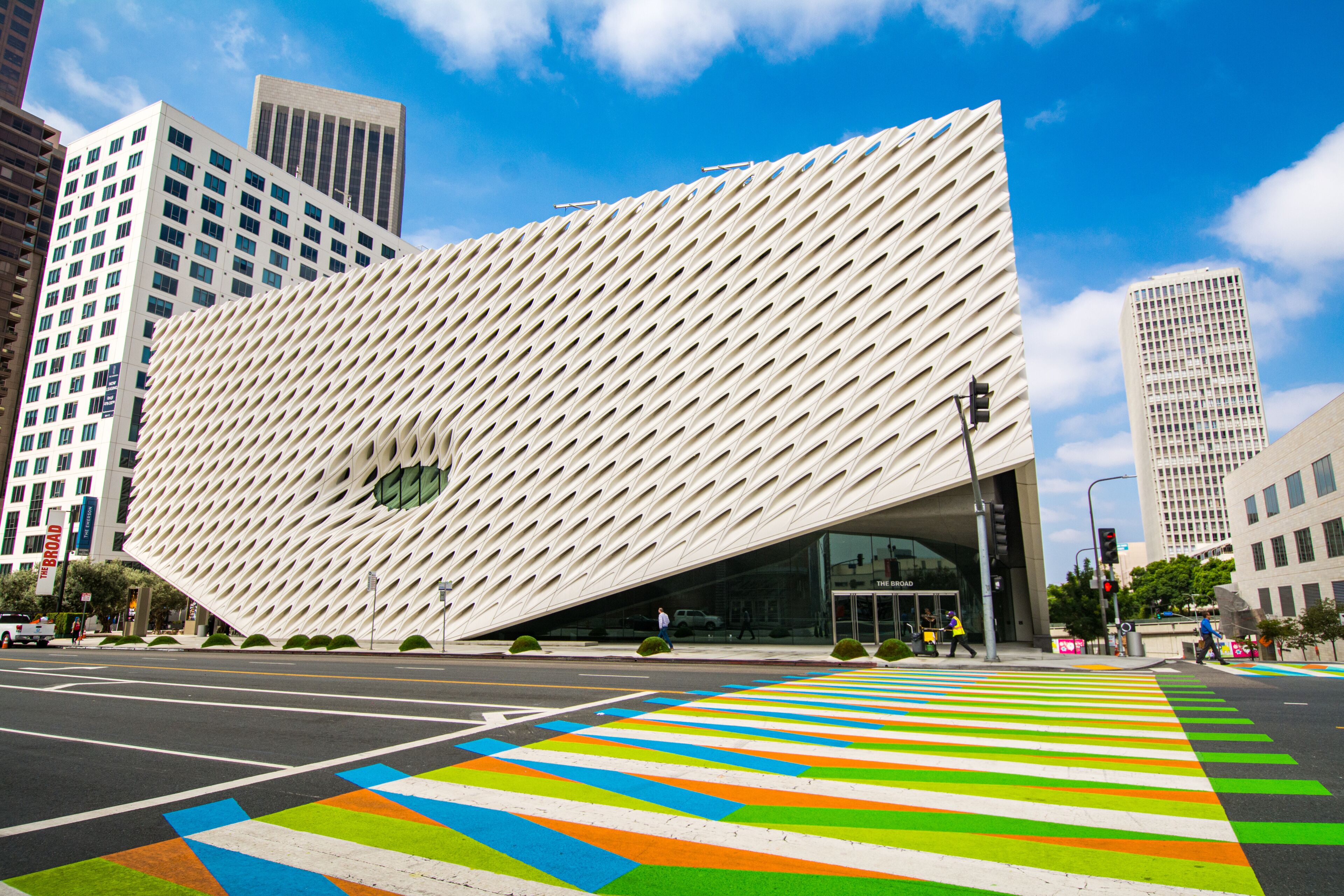 Um edifício moderno com fachada geométrica singular, uma faixa de pedestres com listras coloridas e arranha-céus urbanos ao redor.