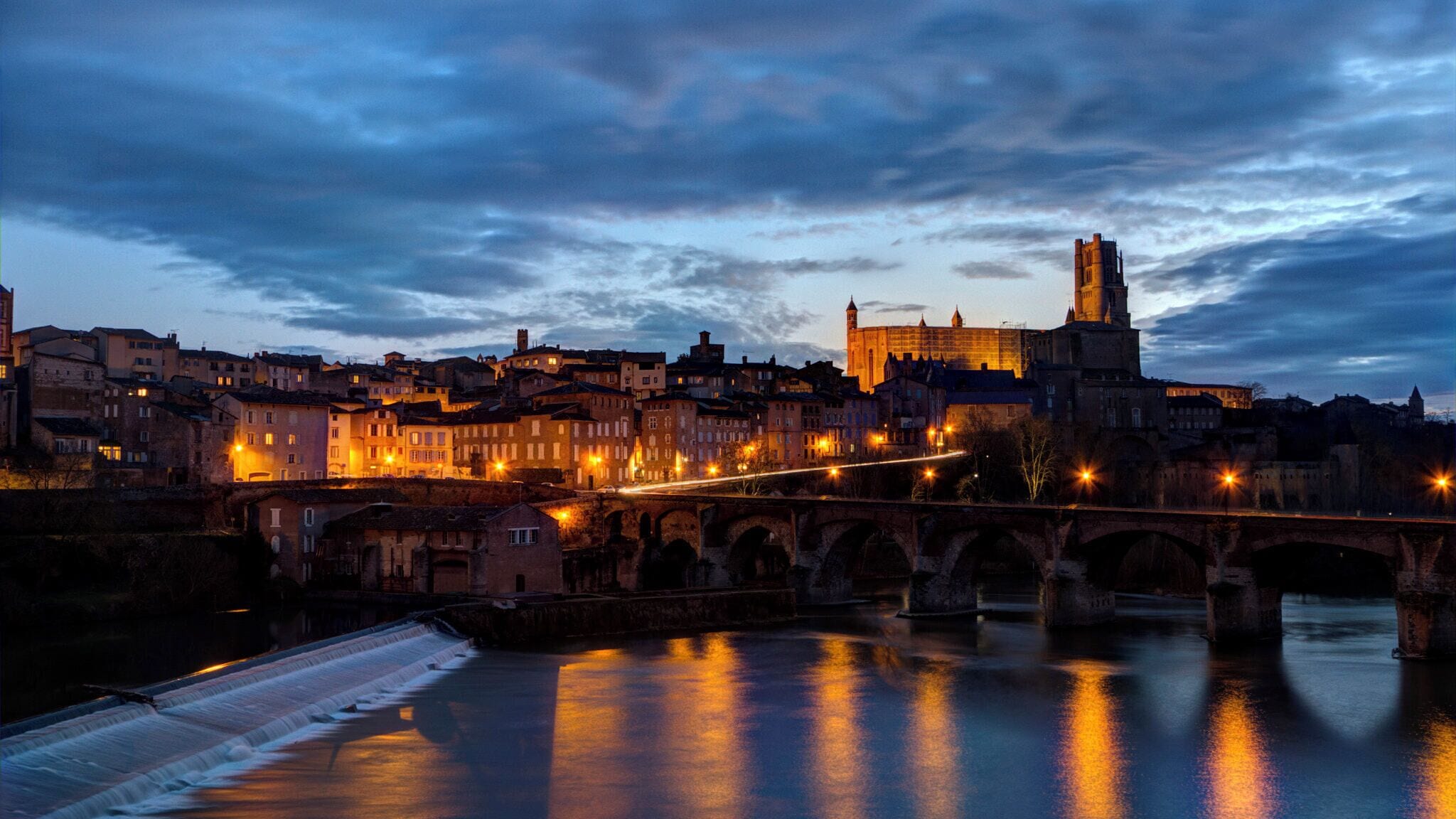 Paisagem urbana ao entardecer, com edifícios iluminados, uma torre imponente e uma ponte sobre o rio.
