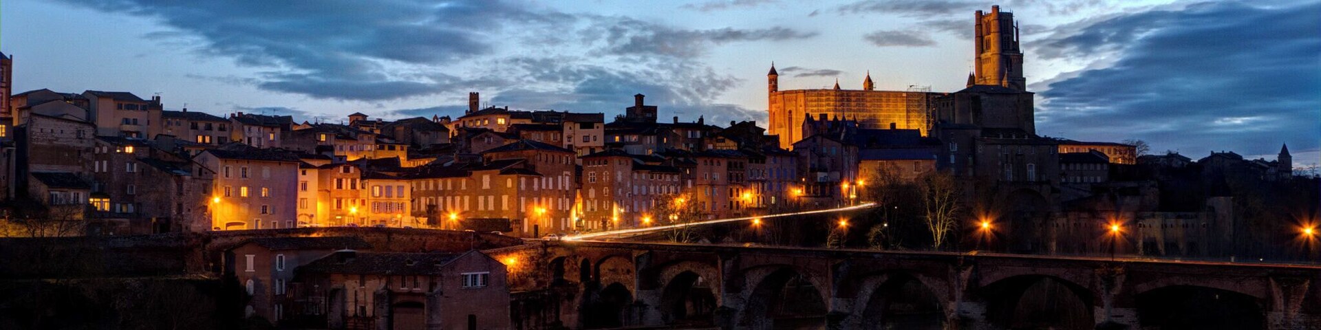 Paisagem urbana ao entardecer, com edifícios iluminados, uma torre imponente e uma ponte sobre o rio.